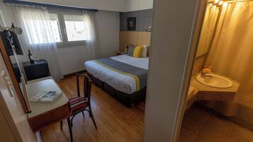 Quarto de hotel com cama, escrivaninha com cadeira, mesinha com revista, janela com cortinas e banheiro com pia e espelho.