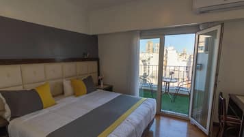 Quarto de hotel com cama, mesa de cabeceira, janela com vista para edifícios e varanda com cadeiras.