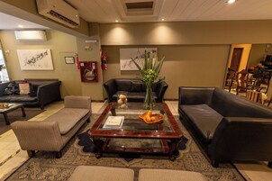 Um hall de hotel com sofás de couro, uma mesa de centro com livros e laranjas, um vaso com flores e um extintor de incêndio.