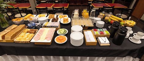 Mesa de buffet com diversos alimentos e bebidas, rodeada por mesas e cadeiras.