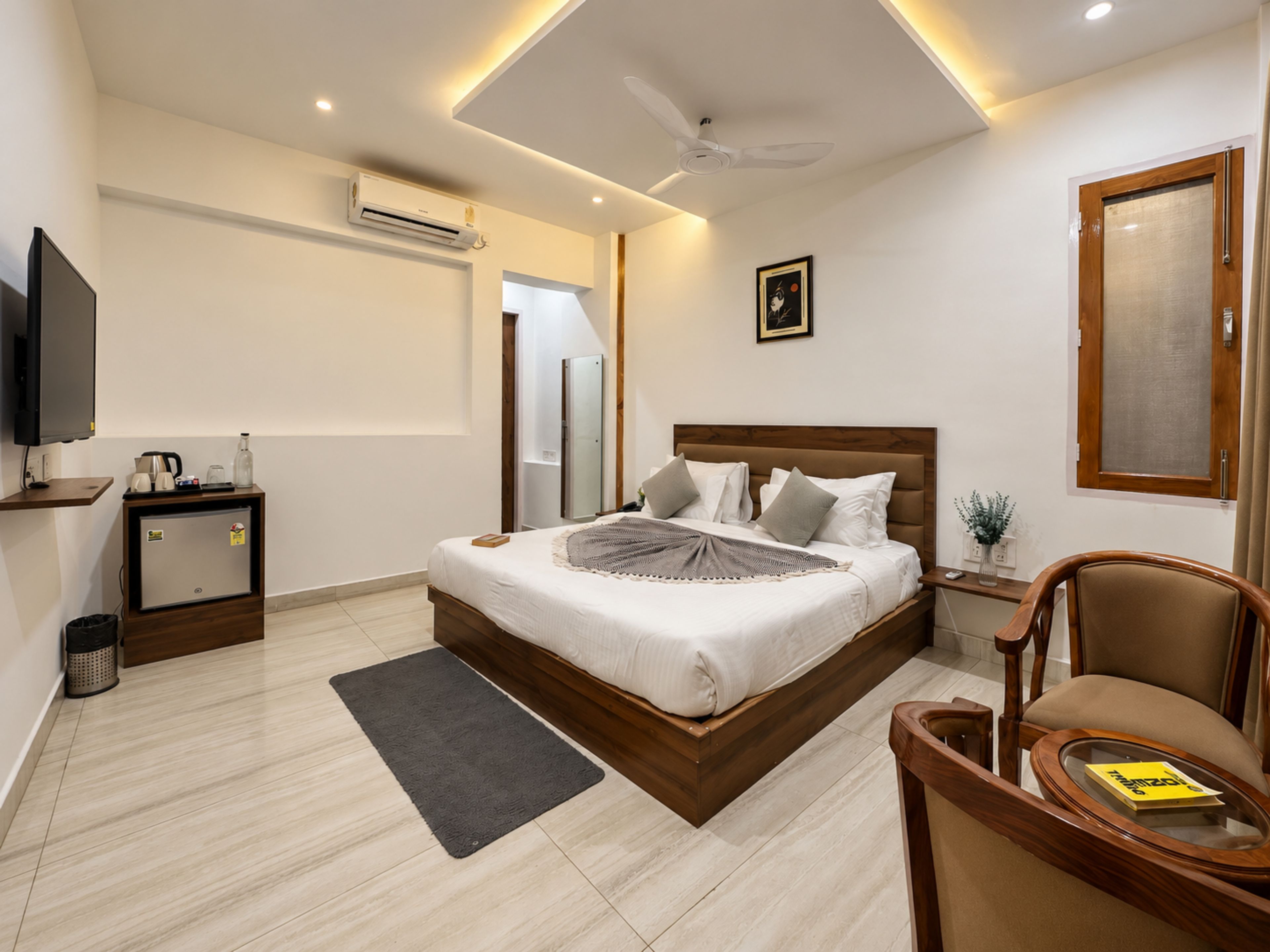 Quarto de hotel com cama, mesa de cabeceira, cadeira, televisão e pequeno frigorífico.