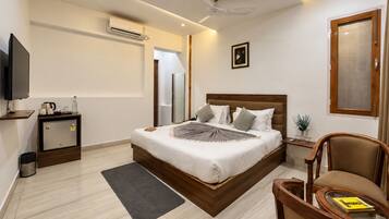 Quarto de hotel com cama, mesa de cabeceira, cadeira, televisão e pequeno frigorífico.
