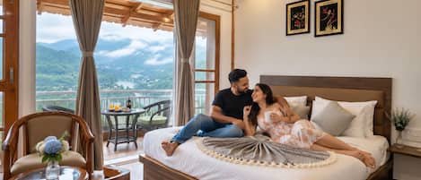Quarto com teto de madeira, cama com roupa de cama branca, vista para montanhas e área de jantar com mesa e cadeiras.