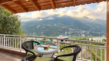 Varanda de madeira com móveis de vime e mesa de vidro, com vista para uma paisagem montanhosa e um lago.