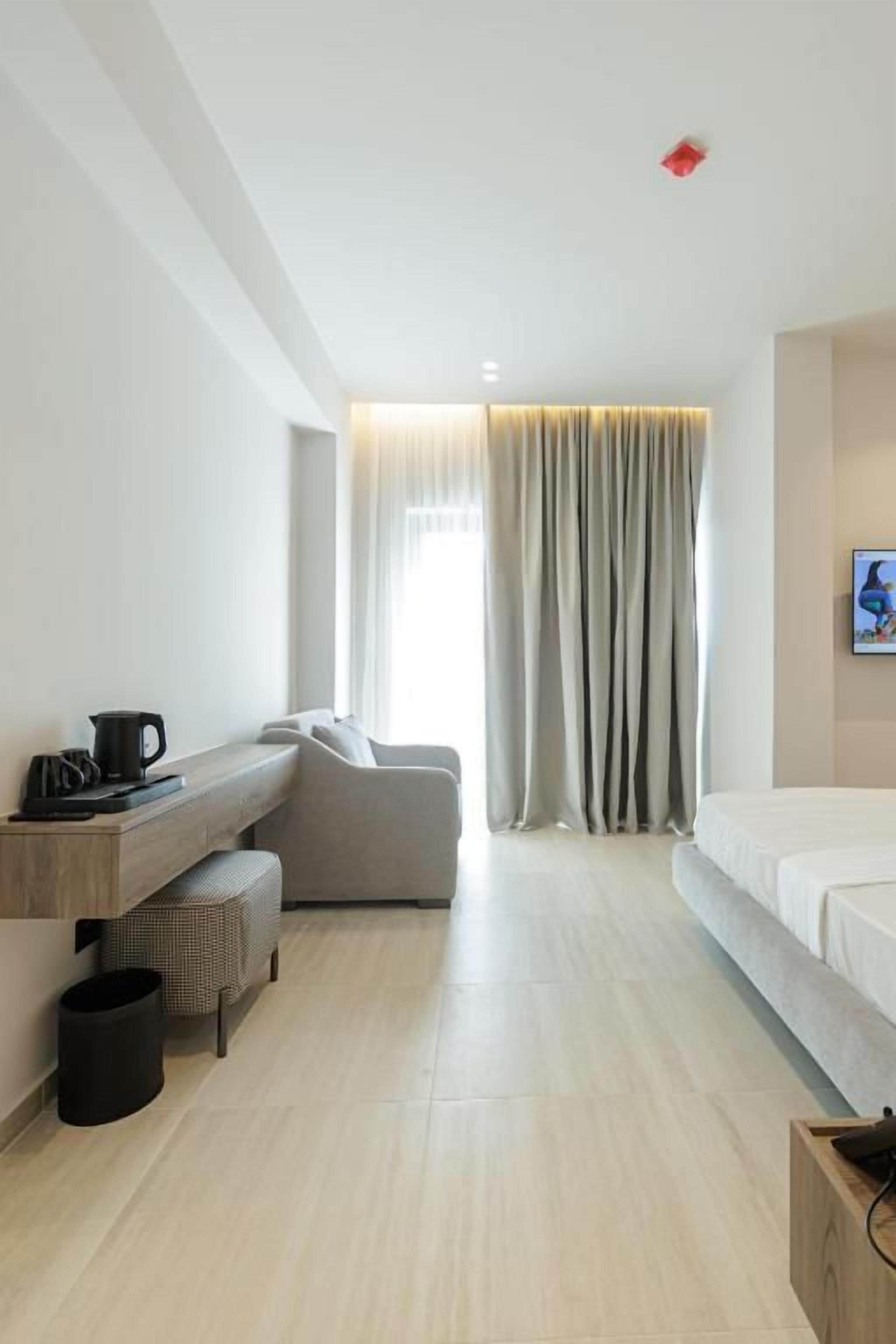 Quarto de hotel moderno com sofá, uma mesinha com chaleira, cama e janela com cortinas.