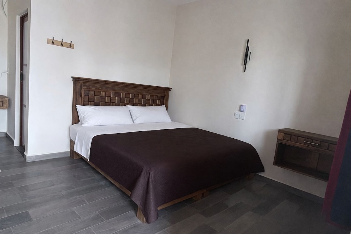 Quarto com cabeceira de madeira, cama com roupa de cama branca e castanha, mesa de cabeceira e luminária de parede.