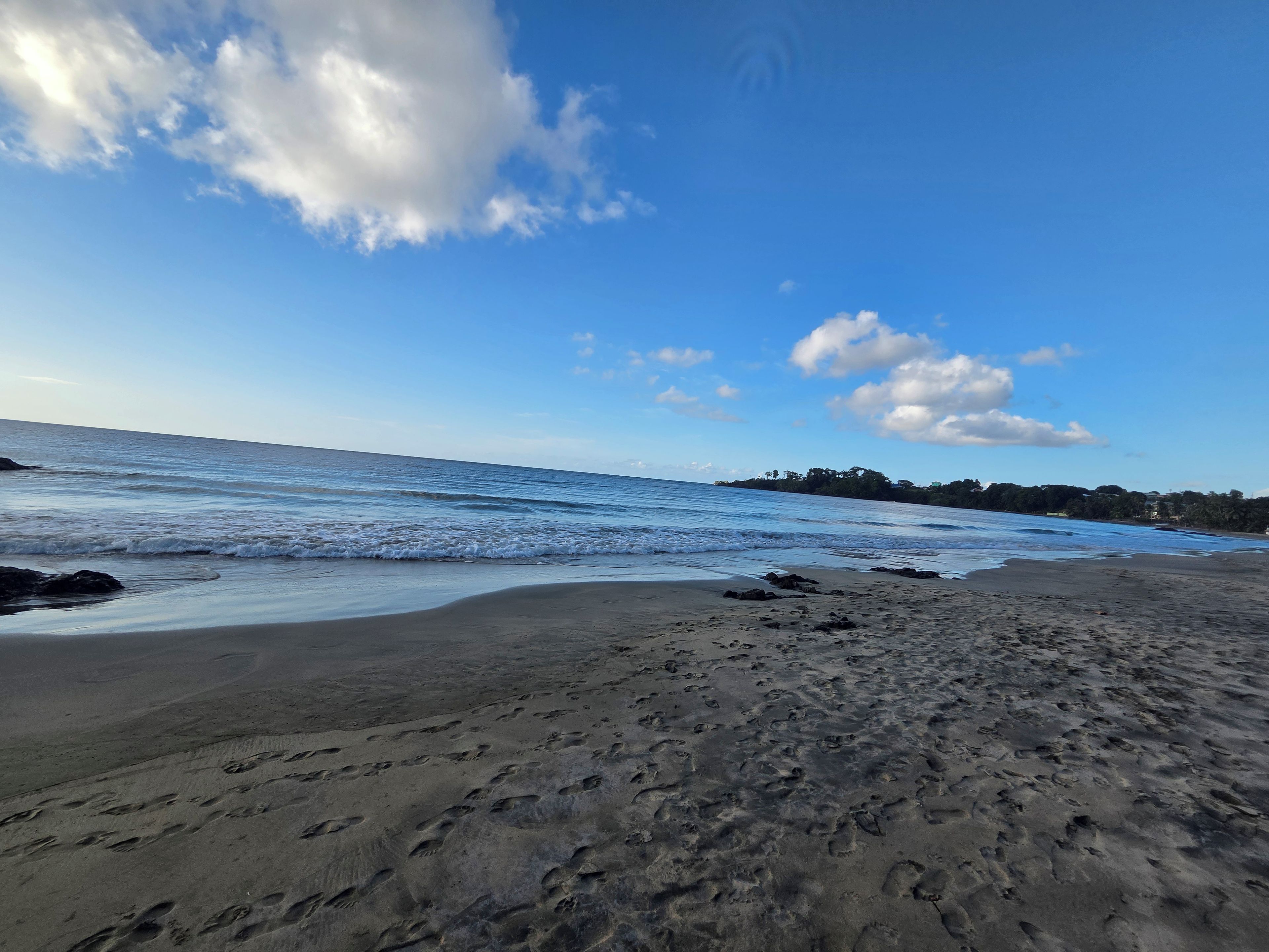 Uma praia com pegadas na areia, céu claro e uma linha de costa distante com árvores.