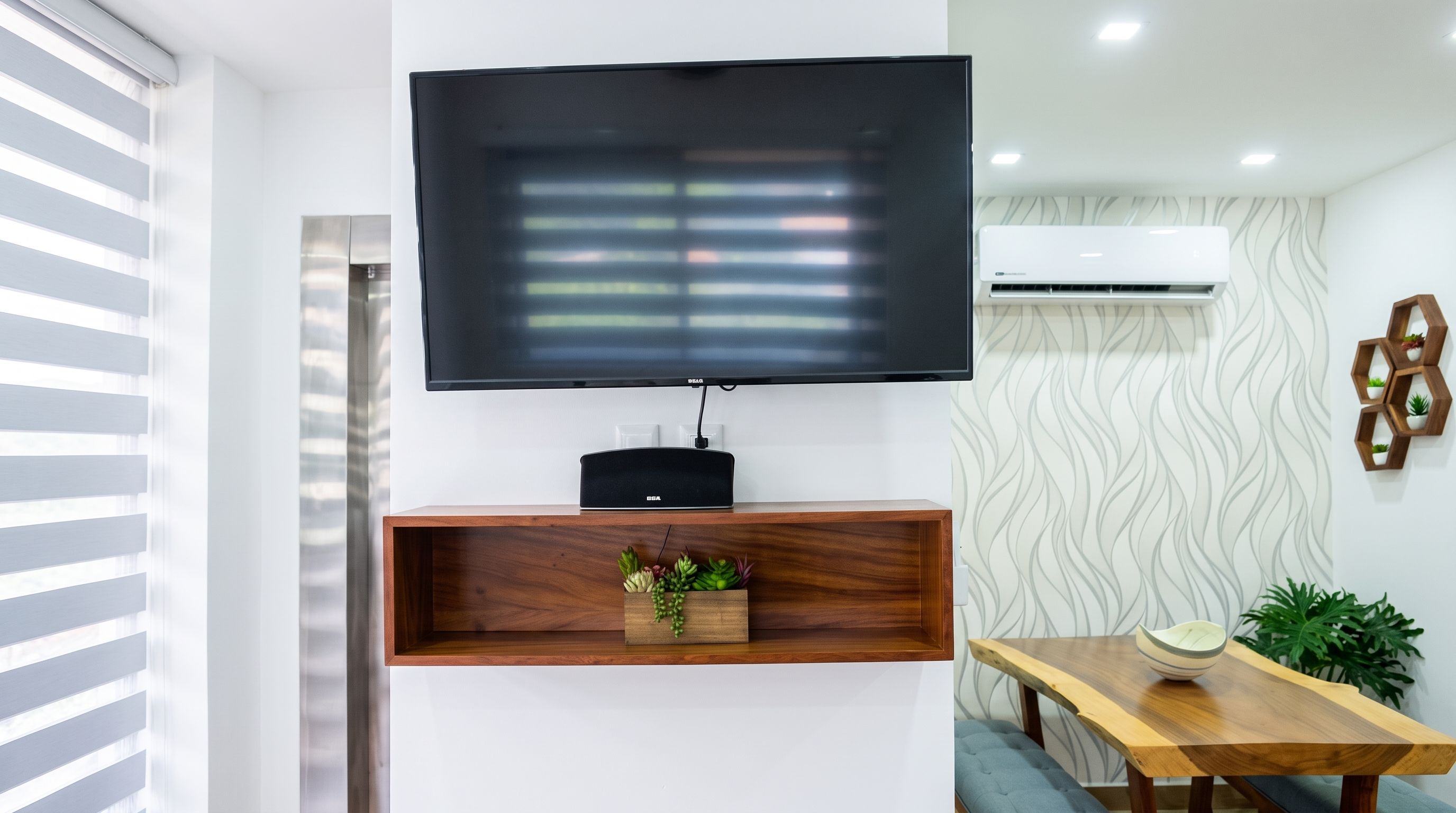 Uma sala de estar moderna com uma televisão de tela plana na parede, uma prateleira de madeira com uma planta e um espaço de jantar com uma mesa e cadeiras de madeira.