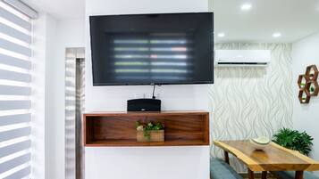 Uma sala de estar moderna com uma televisão de tela plana na parede, uma prateleira de madeira com uma planta e um espaço de jantar com uma mesa e cadeiras de madeira.