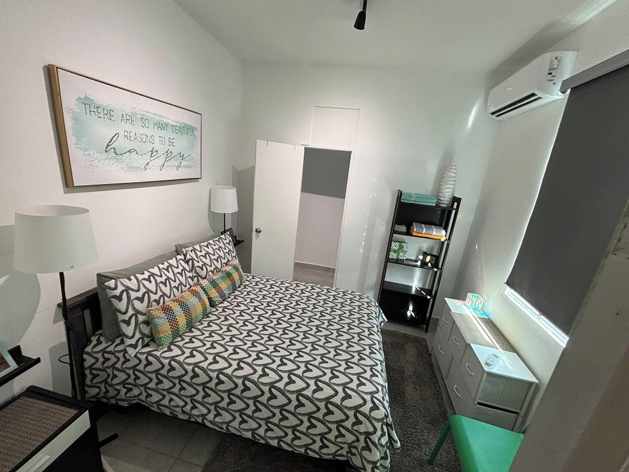 Quarto com cama, luminárias de cabeceira, escrivaninha com cadeira, guarda-roupa e ar condicionado.