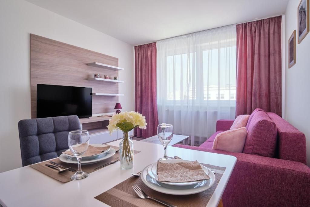 Quarto de hotel com mesa de jantar posta para dois, televisão de tela plana na parede, sofá e janela com cortinas leves.