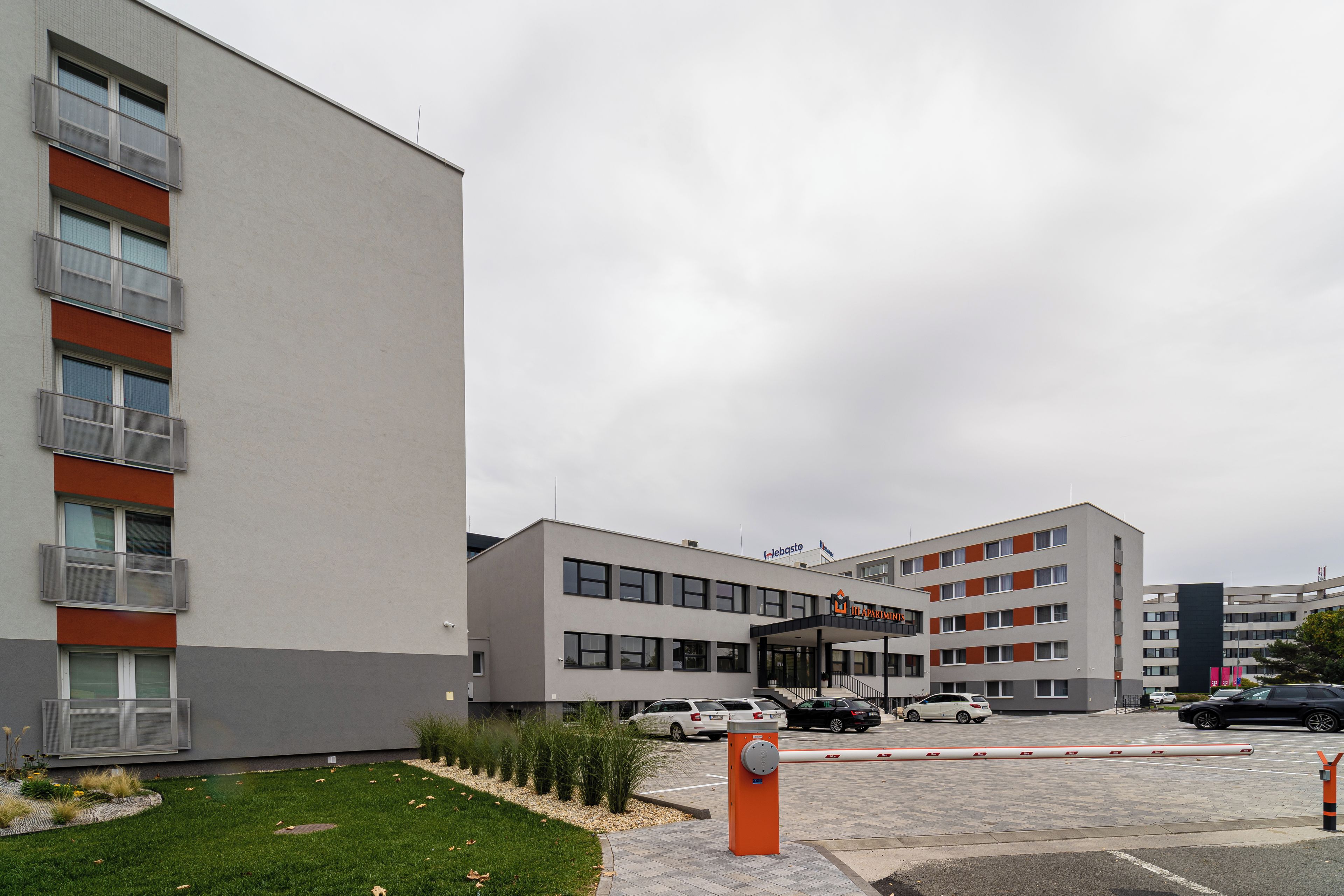 Hotel de vários andares com design moderno, apresentando área de estacionamento com carros e portão de acesso.