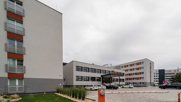 Hotel de vários andares com design moderno, apresentando área de estacionamento com carros e portão de acesso.