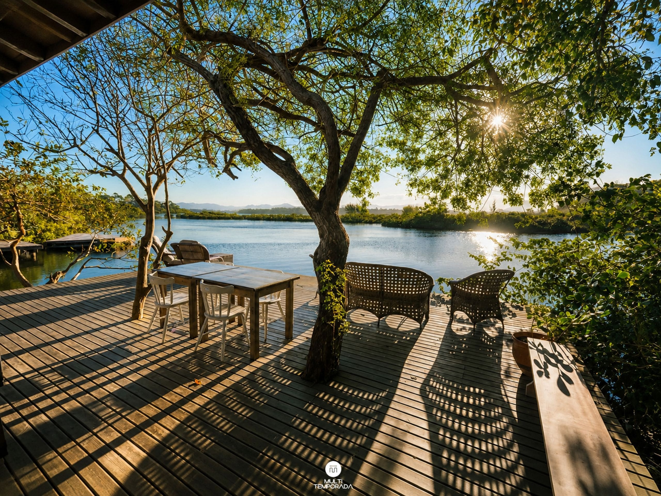 Um deck de madeira com mesa e cadeiras, com vista para um lago calmo, árvores e céu claro.
