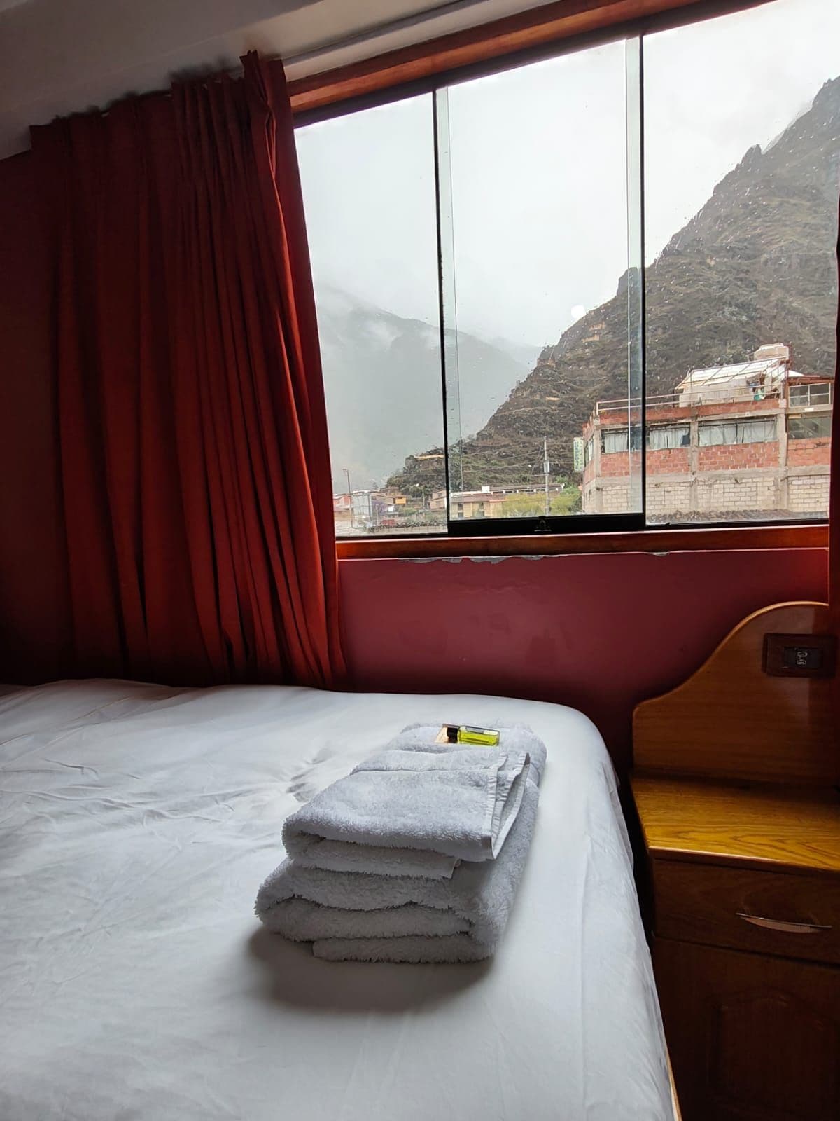 Quarto de hotel com uma cama, janela com vista para as montanhas e mesa de cabeceira de madeira.