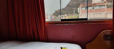 Quarto de hotel com uma cama, janela com vista para as montanhas e mesa de cabeceira de madeira.