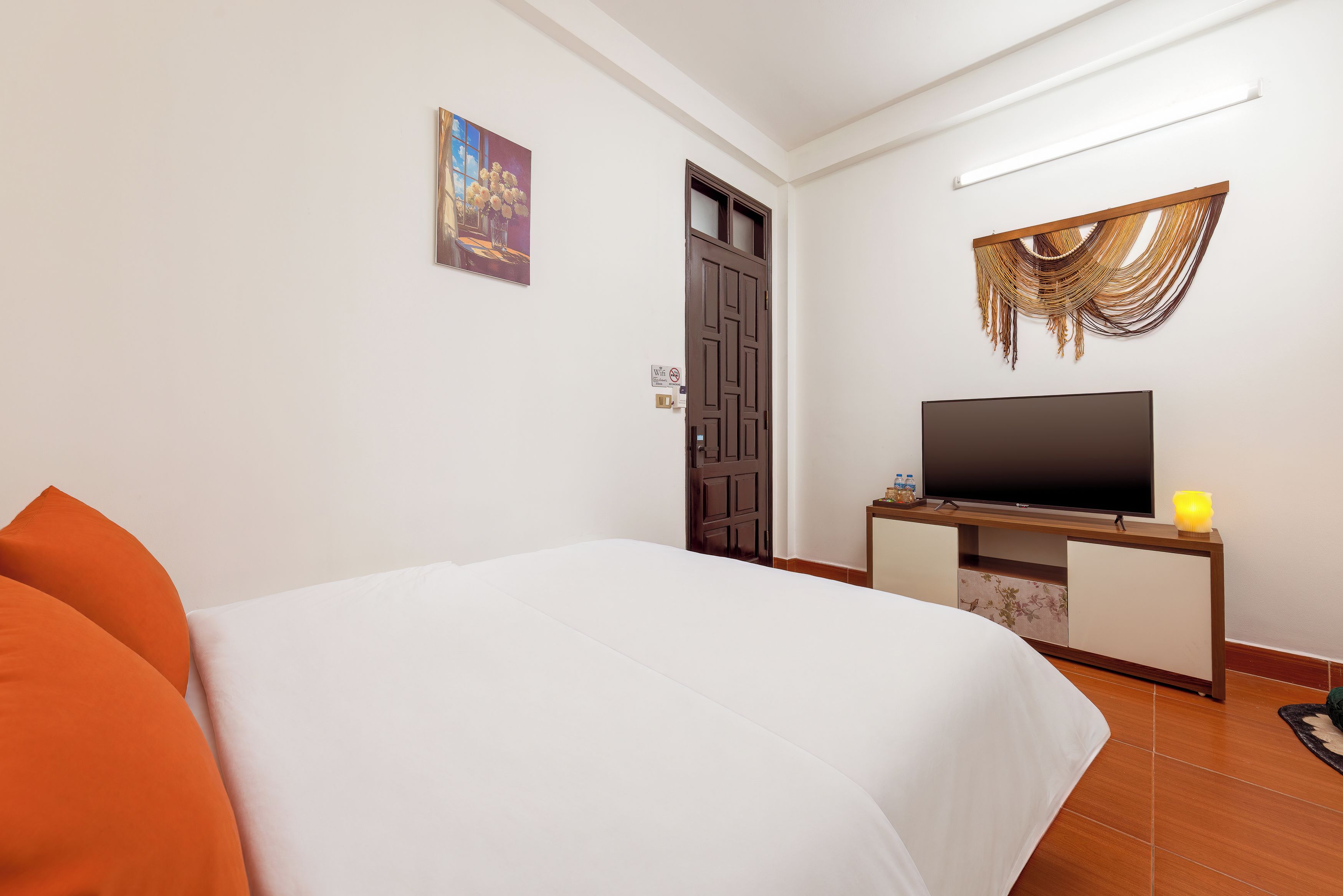 Quarto de hotel com cama, televisão sobre suporte de madeira, porta e luminária fixada na parede.