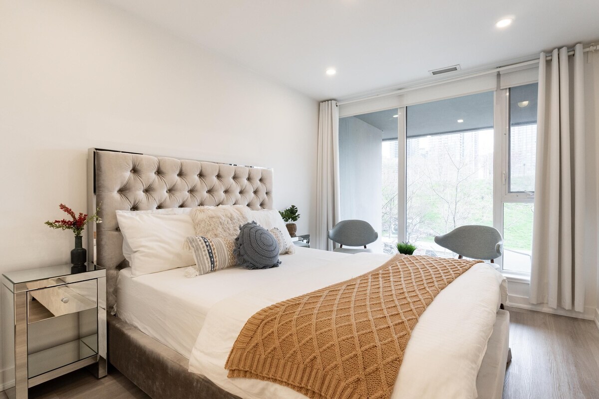 Quarto moderno com uma cama grande, divisória de vidro, uma mesinha e uma cadeira.
