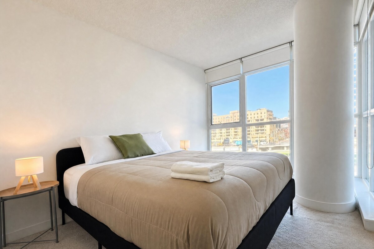 Quarto de hotel moderno com uma cama grande, mesa de cabeceira, luminária e vista da cidade através de amplas janelas.