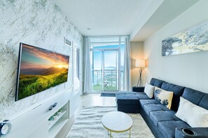 Uma sala de estar moderna com uma televisão de tela grande, um sofá azul escuro, uma mesa de centro redonda e vista para o horizonte urbano pelas amplas janelas.
