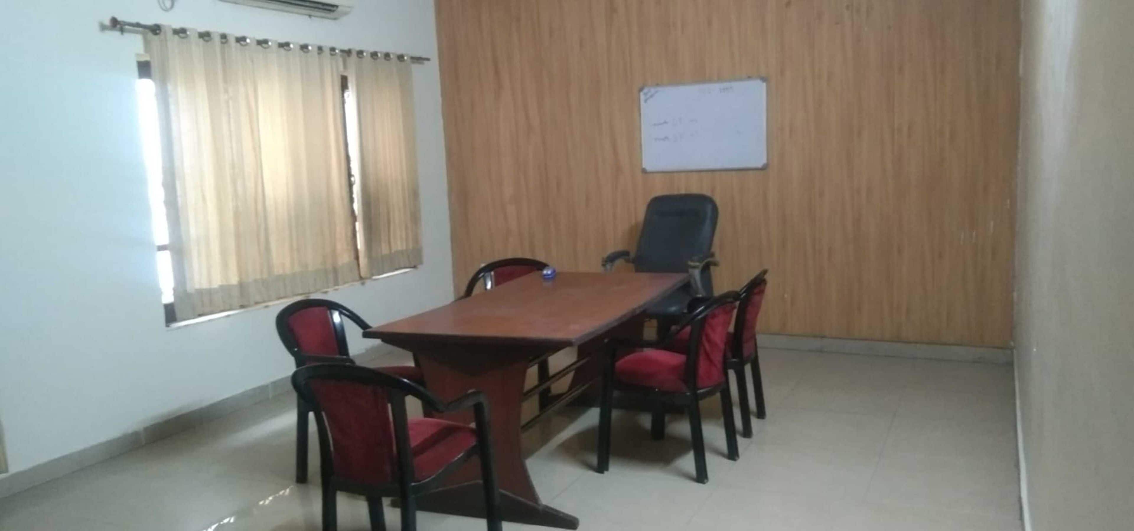 Sala de conferências com mesa de madeira, quatro cadeiras, um quadro branco e uma janela com cortinas.