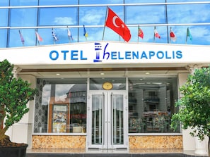 A entrada de um hotel chamado 'OTEL HELENAPOLIS', com fachada de vidro, um conjunto de portas duplas e uma seção inferior revestida de azulejos.