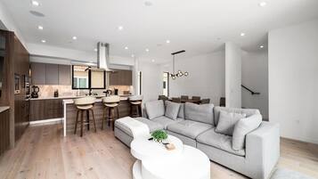 Sala de estar moderna com um sofá cinzento, uma mesa de centro branca e uma área de cozinha com balcão e banquetes.