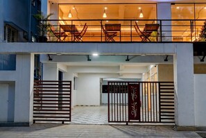 Um edifício de dois andares com portão fechado, varanda com cadeiras e piso xadrez.