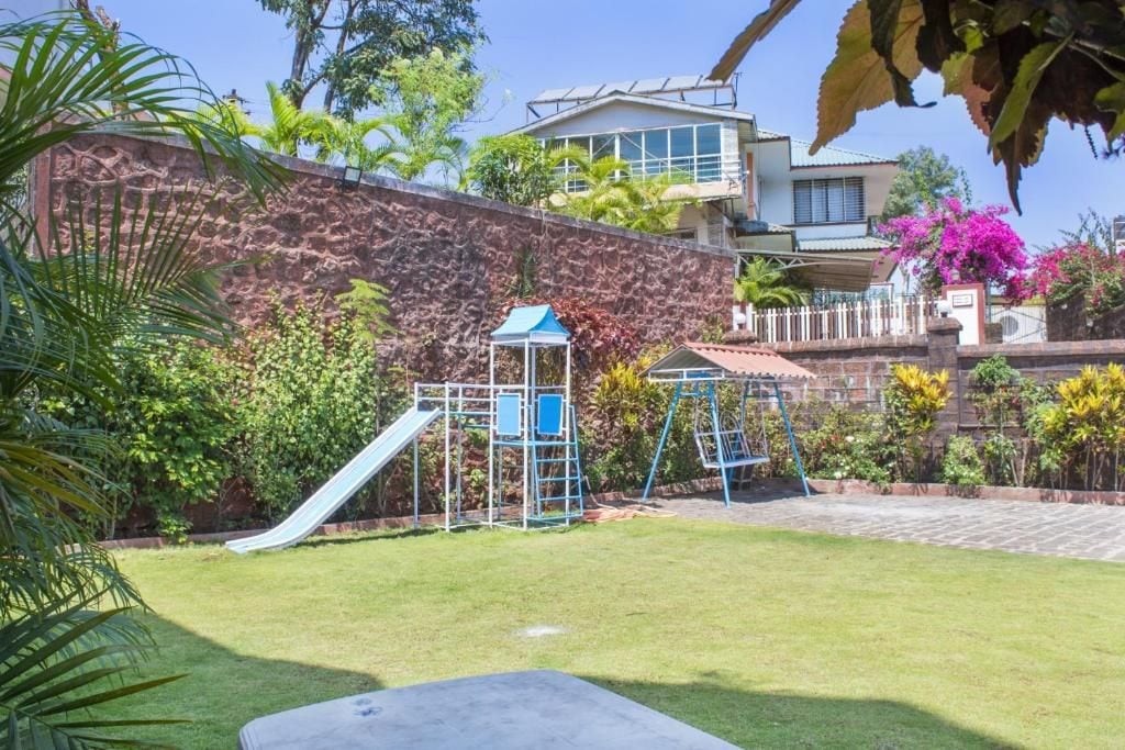 Um bairro residencial com quintal gramado, estrutura de recreação infantil e uma casa com muro de pedra e varanda.