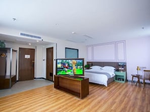 Quarto de hotel com piso de madeira, cama com roupa de cama branca, televisão sobre um suporte de madeira e mesa de cabeceira com abajur.