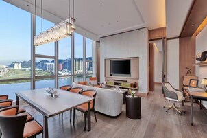 Quarto de hotel moderno com televisão grande, área de estar e vista para a paisagem urbana.