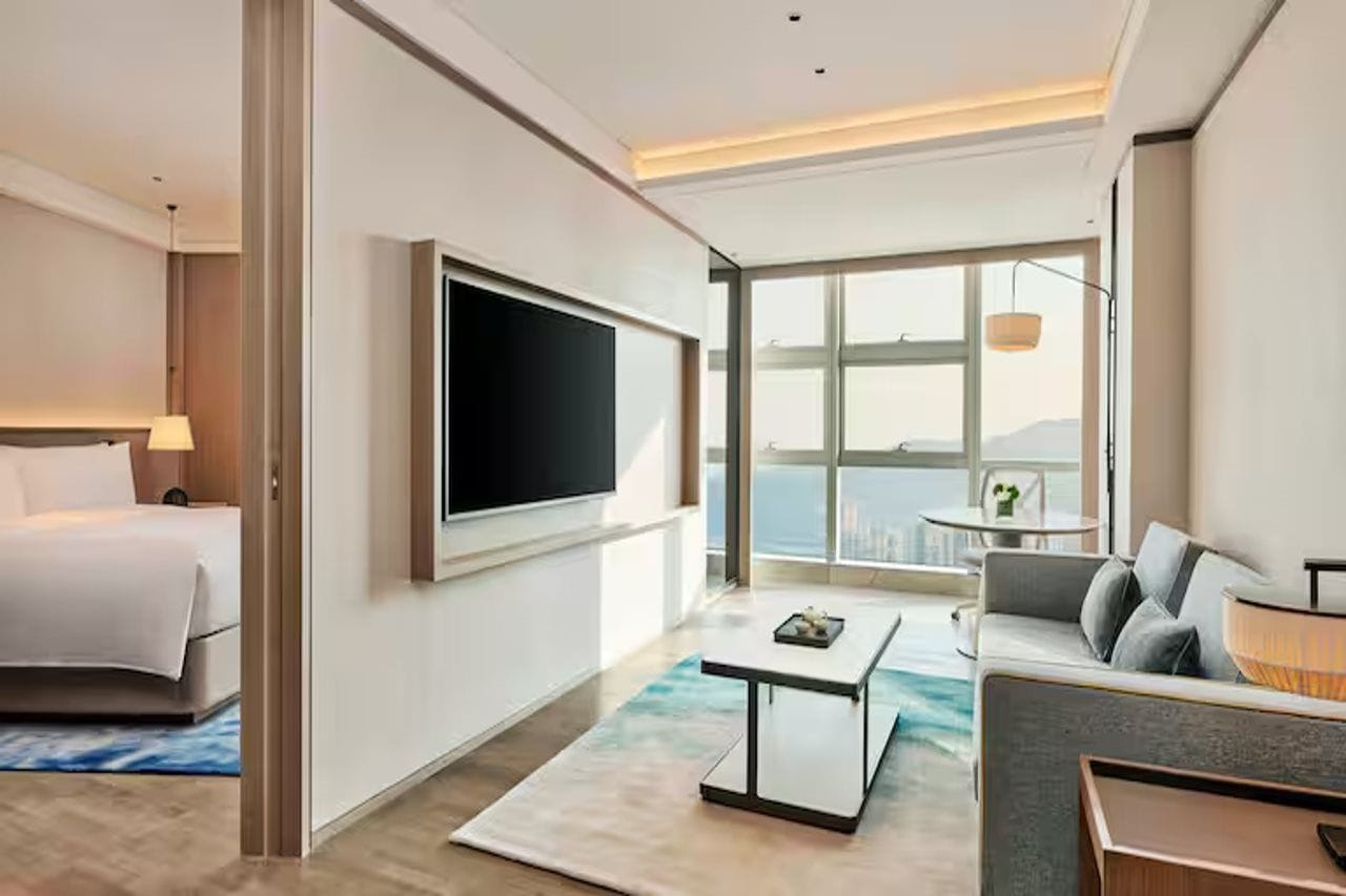 Quarto de hotel moderno com cama, televisão de tela plana, sofá, mesa de centro e uma janela grande com vista.