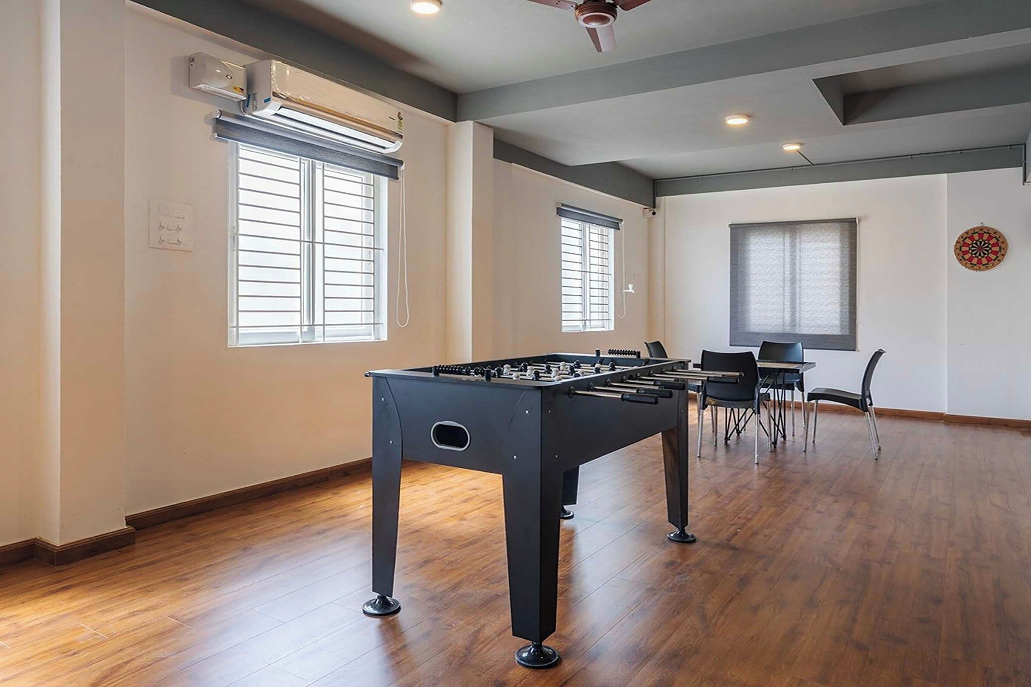 Quarto com mesa de futebol de mesa, duas janelas com persianas, ventilador de teto e alvo de dardos.