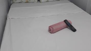Uma cama com lençol branco, toalha rosa enrolada, controle remoto e dois travesseiros estampados com flores.