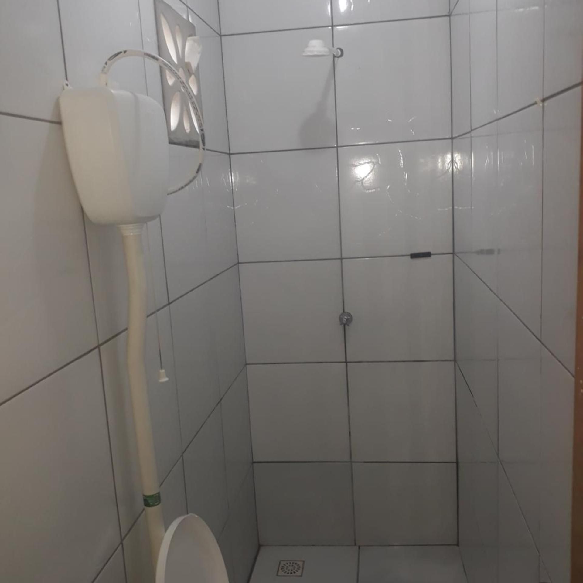 Casa de banho com azulejos brancos, sanita, chuveiro e suporte de papel higiénico fixado na parede.