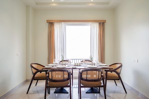 Uma área de jantar com mesa posta para seis pessoas, rodeada por cadeiras de vime, uma janela grande com cortinas e vista para o exterior.
