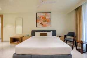 Quarto de hotel com uma cama, uma cadeira, uma mesinha de cabeceira, um criado-mudo, um telefone e um quadro na parede.