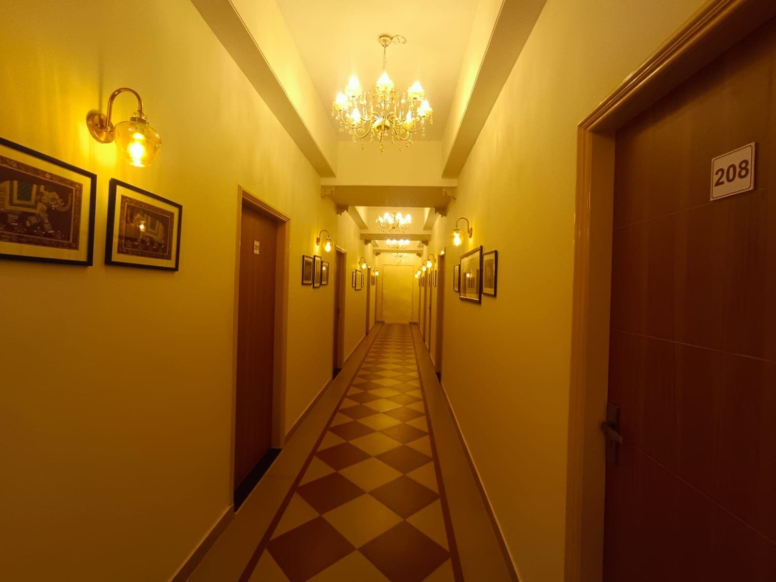 Um corredor de hotel com piso xadrez, luminárias fixadas na parede e um lustre.