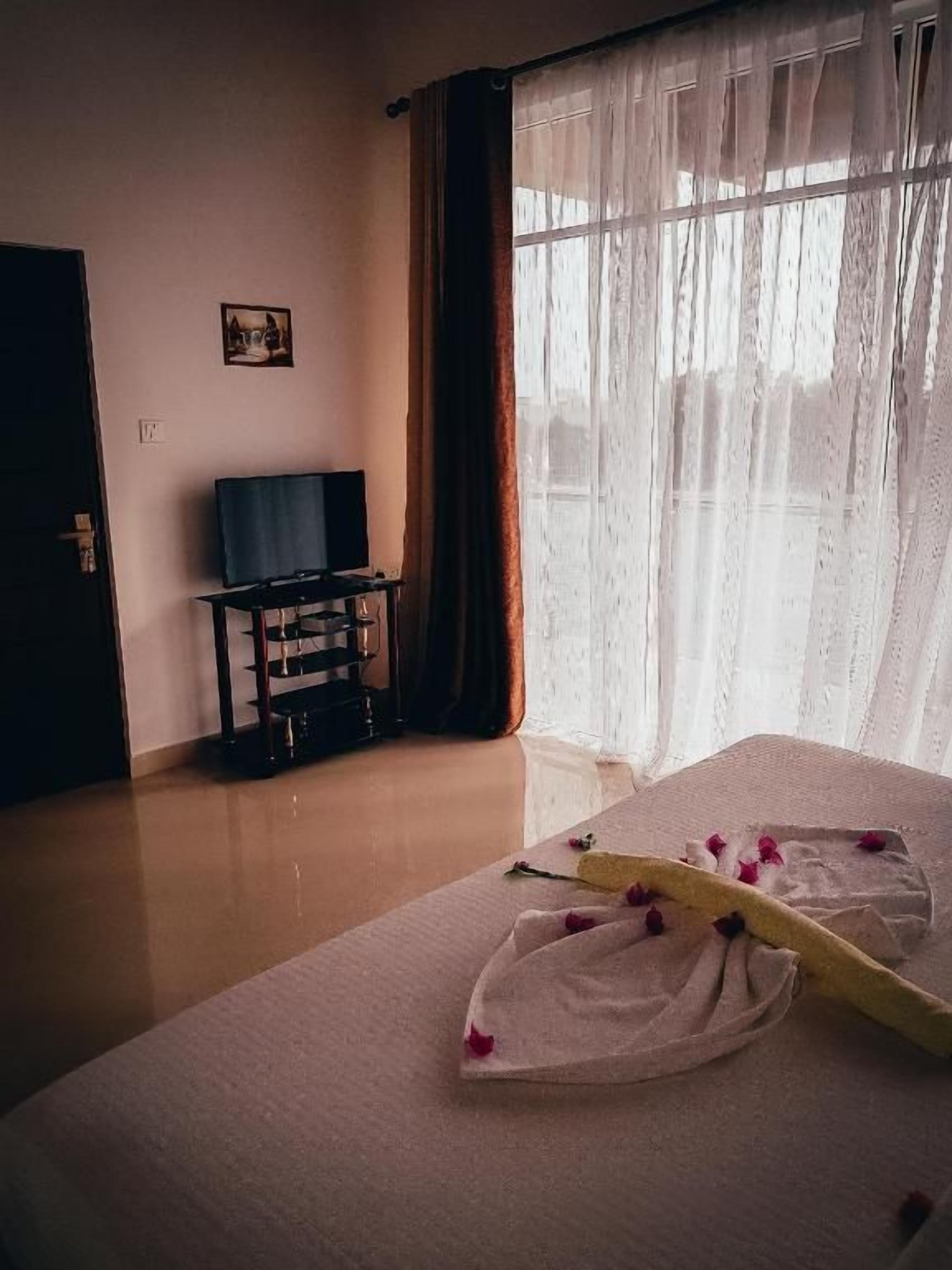 Quarto de hotel com cama, televisão em suporte, janela com cortinas leves e porta.