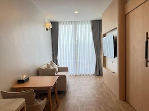 Quarto de hotel com sofá, mesa de jantar, cadeiras, televisão de tela plana fixada na parede e porta de correr para uma varanda.