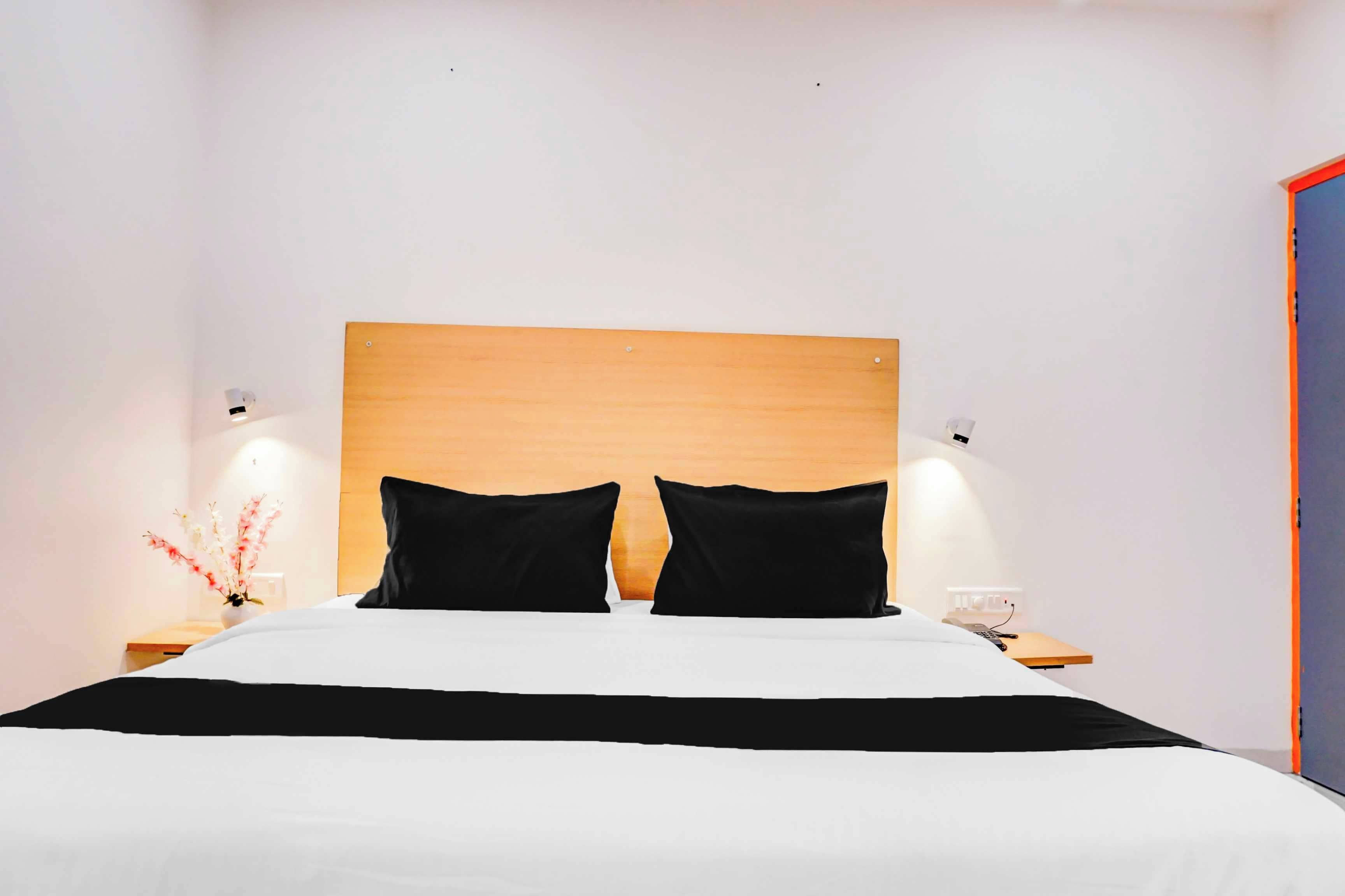 Quarto de hotel com cabeceira de madeira, duas almofadas pretas, colcha branca e mesa de cabeceira com telefone e um vaso de flores.