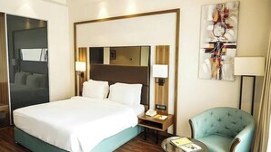 Quarto de hotel com uma cama grande, mesas de cabeceira, uma cadeira, uma pequena mesa e um quadro na parede.