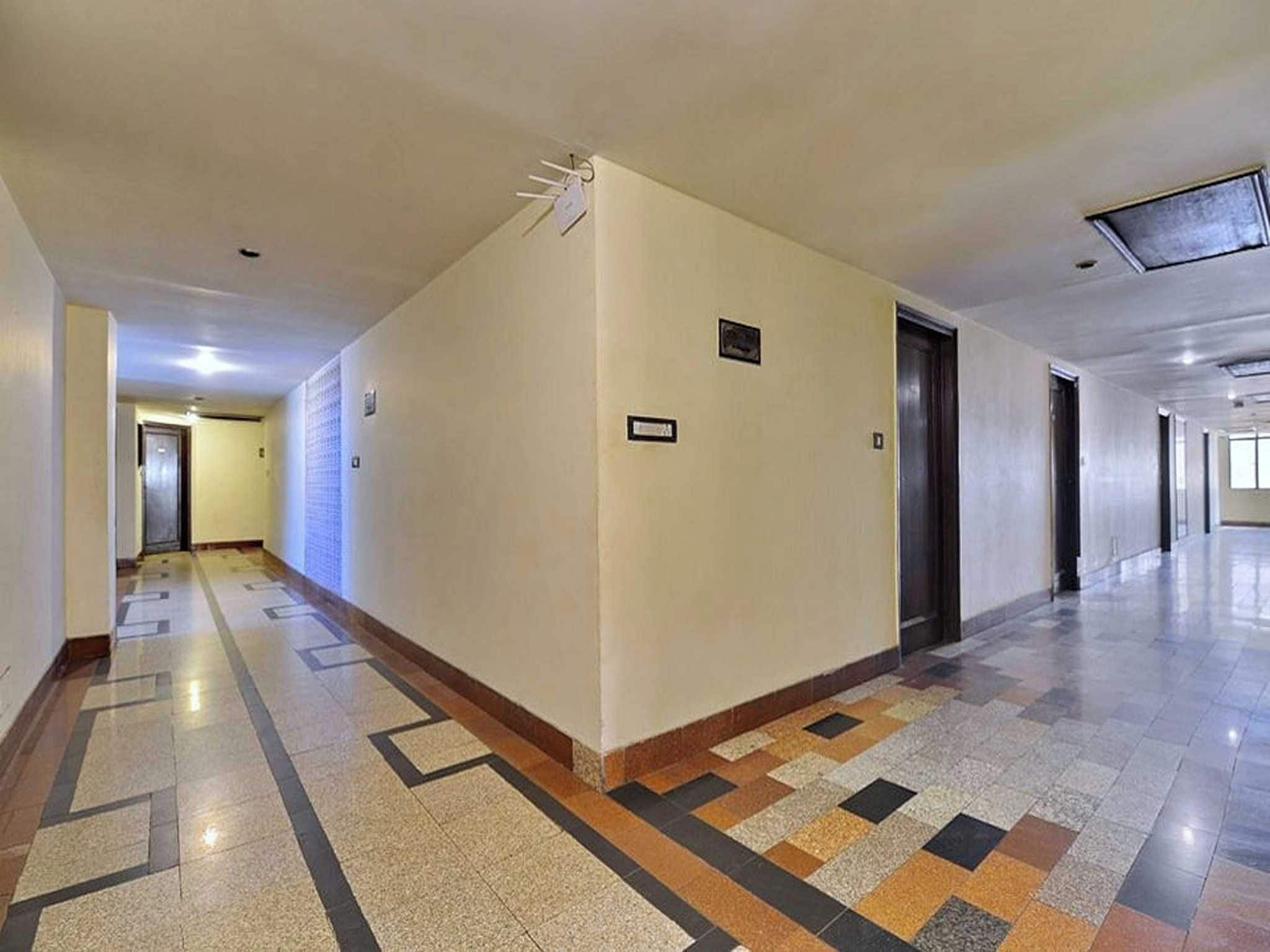 Um corredor longo e estreito com piso estampado, portas de um lado e parede lisa do outro.
