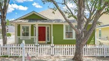 Uma casa pequena, térrea, com fachada verde, porta vermelha e cerca branca de piquete.