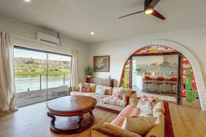 Uma sala de estar ampla com uma grande janela com vista para a água, um ventilador de teto e um espaço de jantar com banquetas.