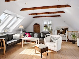 Uma sala de estar no estilo loft, com teto inclinado, vigas de madeira, um sofá de couro preto, uma poltrona branca e uma mesa de centro de madeira.