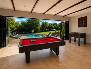 Mesa de bilhar com superfície de feltro vermelho, mesa de futebol de mesa e vista para uma piscina e vegetação lá fora.