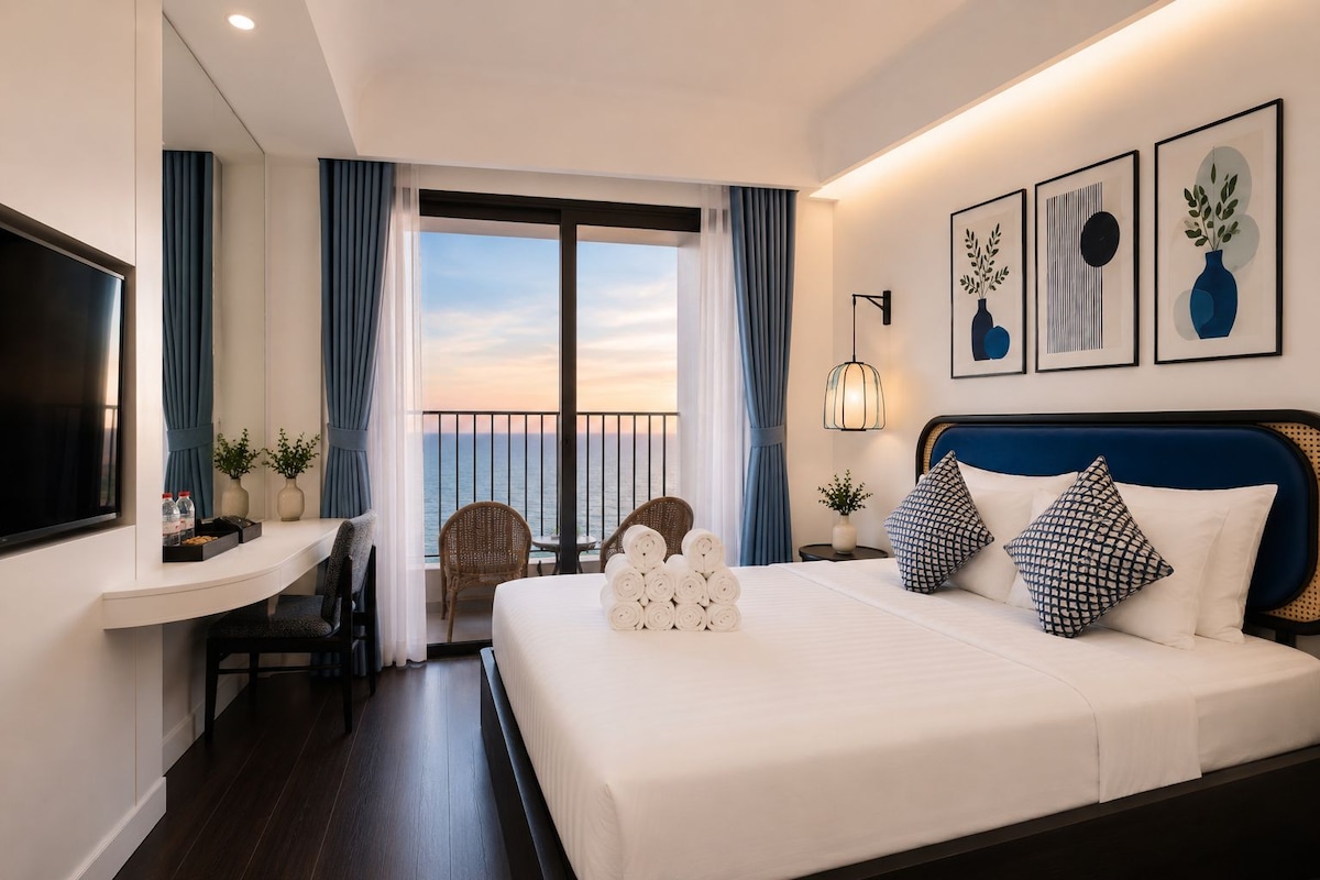 Quarto de hotel com cama, escrivaninha, cadeira, televisão e vista para o mar.