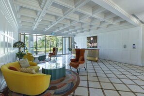 Um hall espaçoso com pé-direito alto, uma janela ampla com vista para o exterior, um balcão de recepção e uma área de estar com uma poltrona amarela e uma mesa redonda.