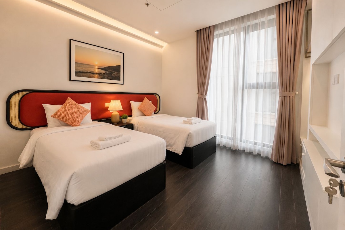 Quarto de hotel com duas camas, uma mesinha de cabeceira com abajur, um quadro pendurado na parede e uma janela grande com cortinas.